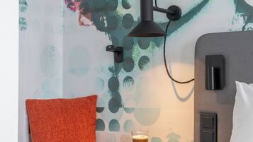 Quarto de hotel com design moderno, com uma cama e cabeceira cinza, um travesseiro vermelho, uma mesinha redonda com luminária e uma parede com uma pintura abstrata colorida.