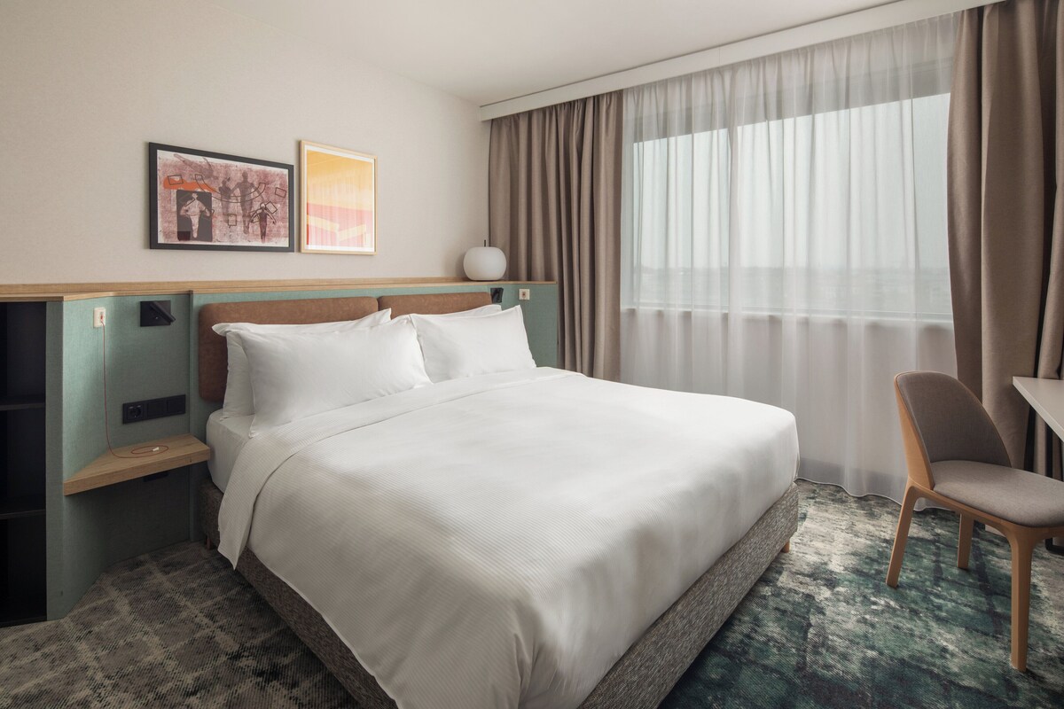 Quarto de hotel com uma cama grande, uma escrivaninha e uma cadeira. Há duas molduras com quadros na parede.