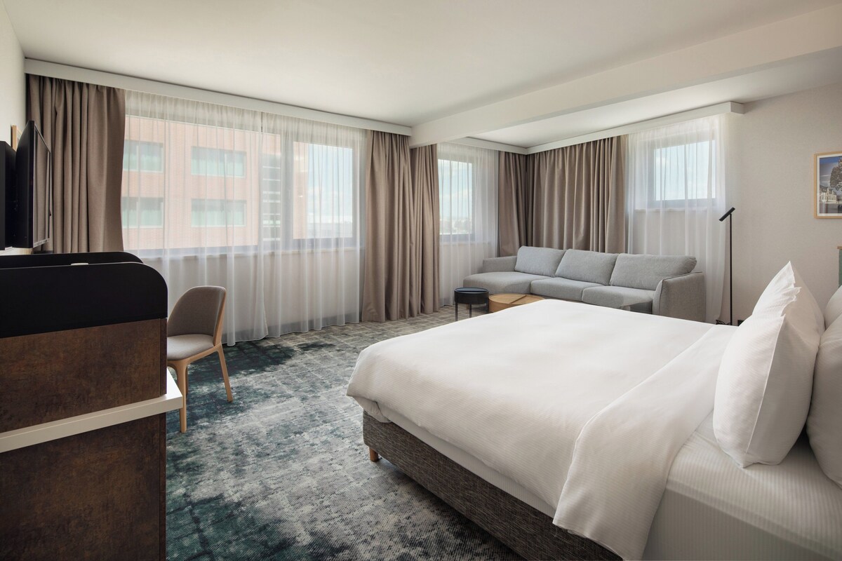 Quarto de hotel com uma cama grande, um sofá, uma cadeira, uma escrivaninha e uma televisão.