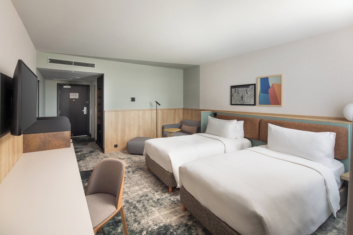 Quarto de hotel com duas camas, uma escrivaninha, uma cadeira, uma televisão e obras de arte na parede.