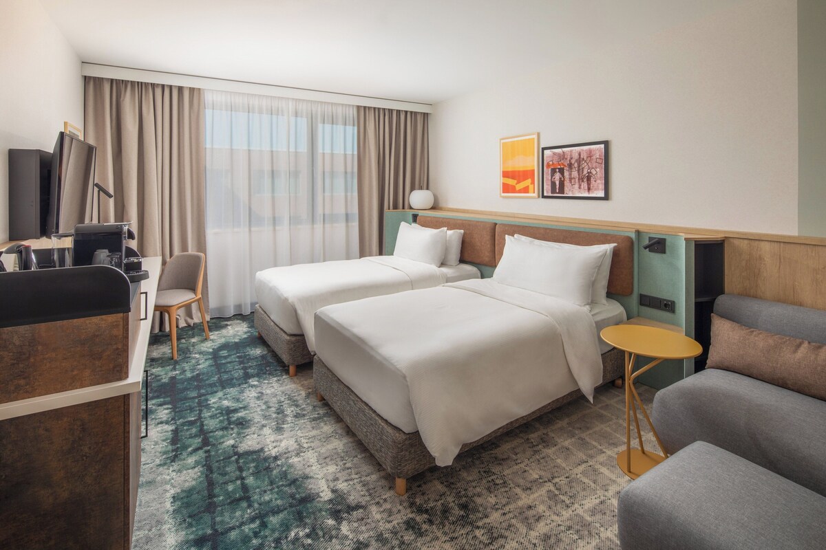 Quarto de hotel com duas camas, uma escrivaninha com computador, uma cadeira, uma mesinha e uma janela grande com cortinas.