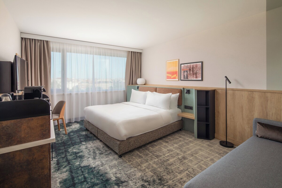 Quarto de hotel com uma cama grande, uma escrivaninha com computador, uma cadeira e uma televisão.