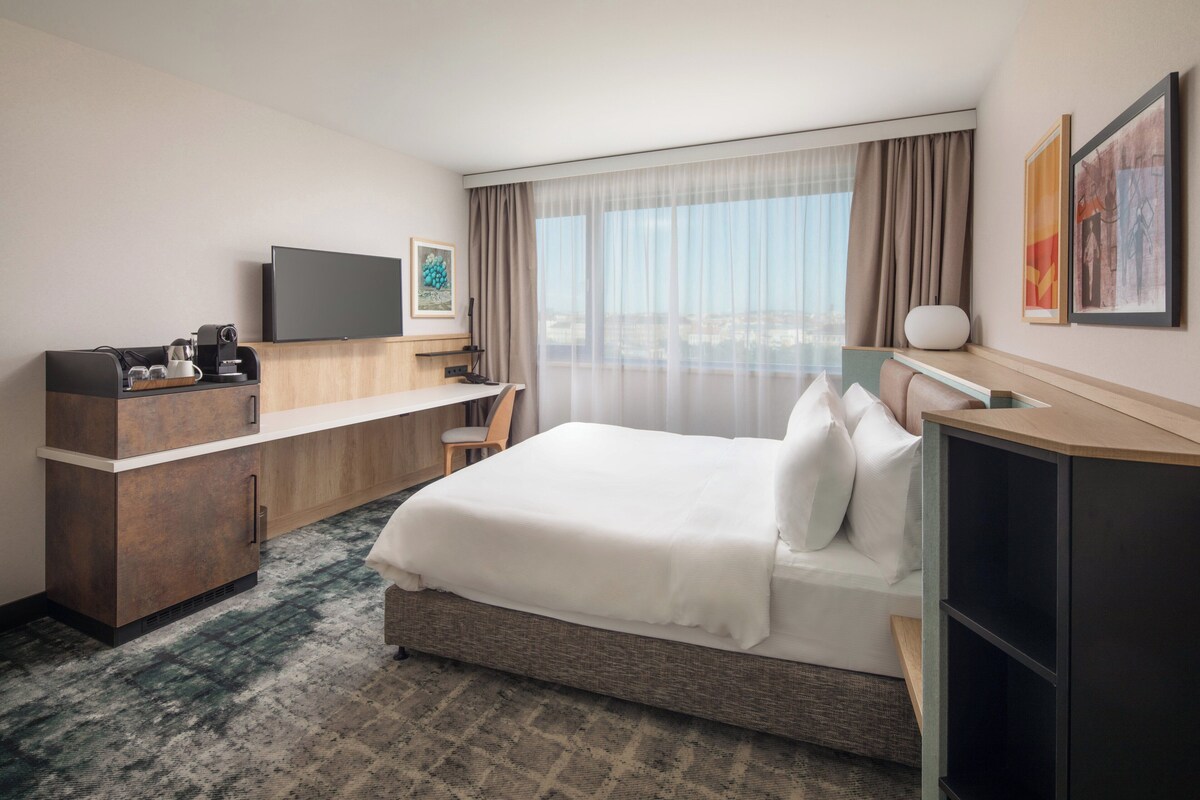 Quarto de hotel com cama, escrivaninha, televisão e janela com cortinas.