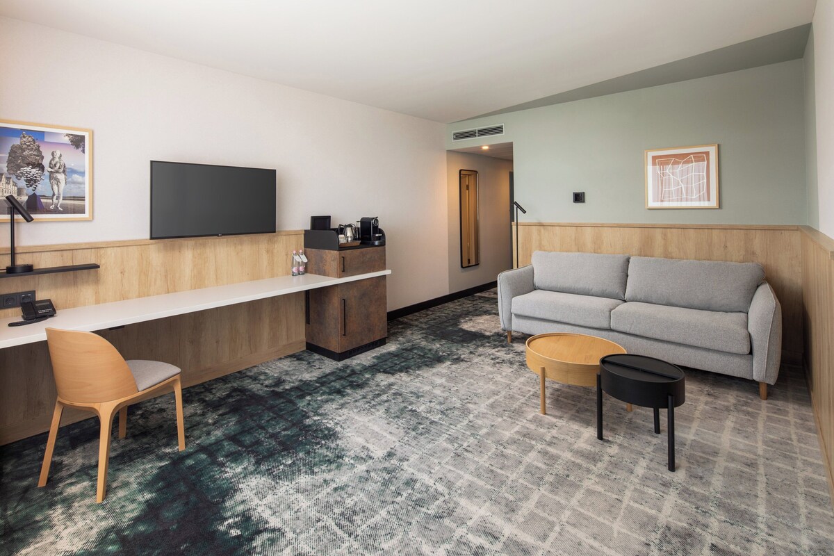 Quarto de hotel moderno com sofá, escrivaninha, cadeira, mesinha de centro e obras de arte nas paredes.