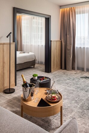 Quarto de hotel com uma cama, mesa de cabeceira com champanhe, copos, bandeja com petiscos e vista para o exterior através de cortinas translúcidas.