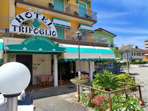 Um hotel chamado "HOTEL TRIFOGLIO" com toldo verde e branco, mesas ao ar livre e um jardim de flores.
