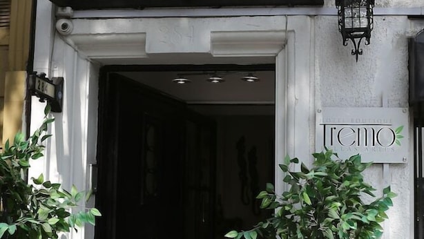 Entrada de hotel com uma porta preta, uma placa acima da porta e duas plantas em vasos de cada lado.