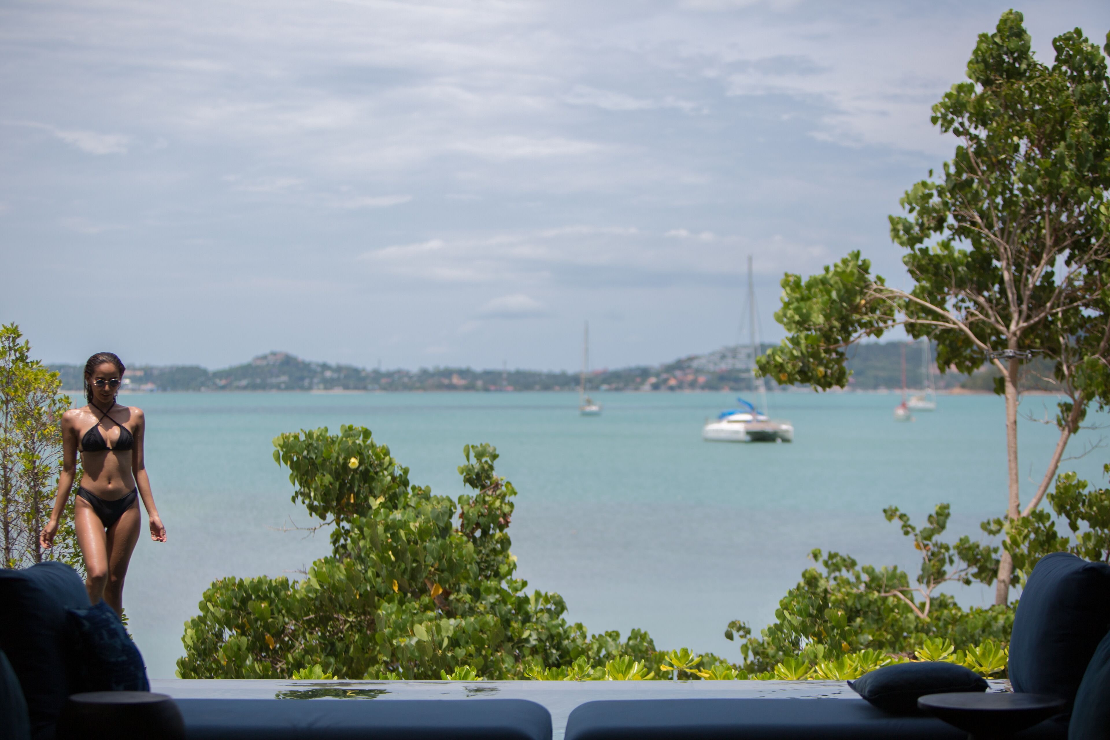 Vista de uma área de piscina com espreguiçadeiras, uma pessoa de fato de banho e um corpo d'água com barcos ao longe.