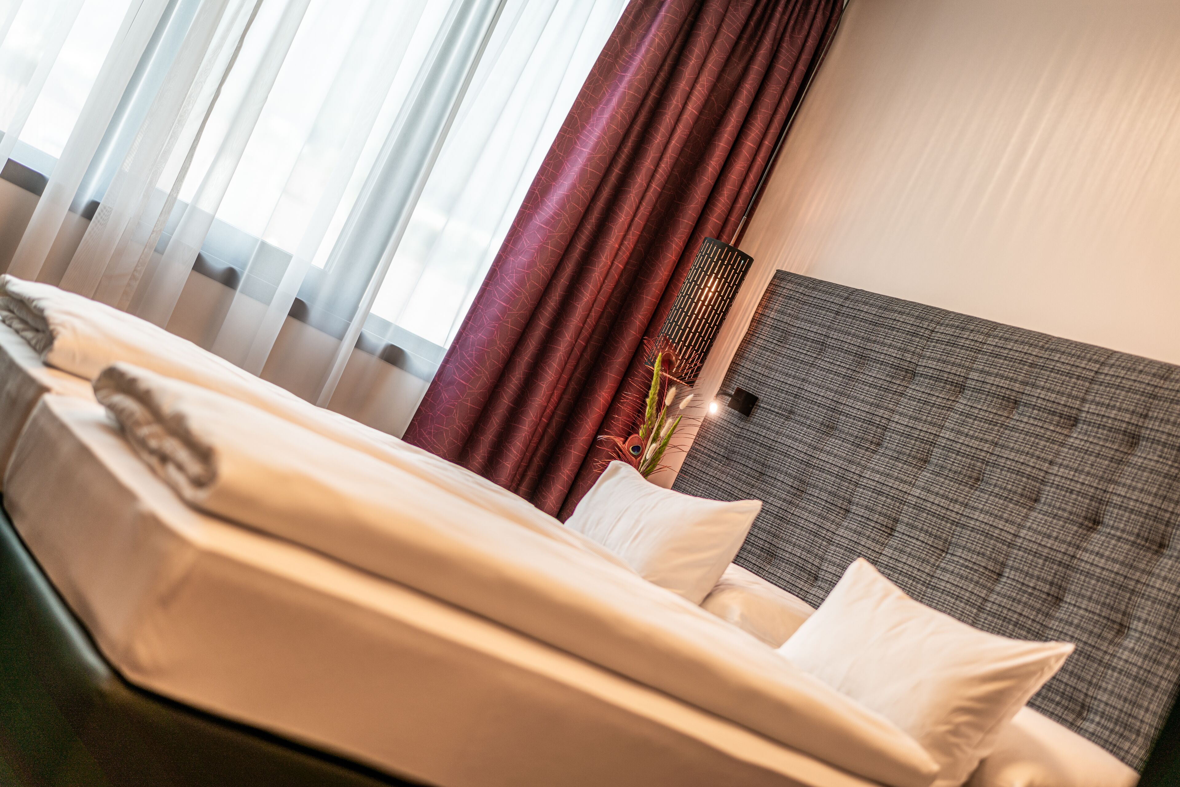 Quarto de hotel moderno com uma cama grande, cabeceira e janela com cortinas translúcidas.