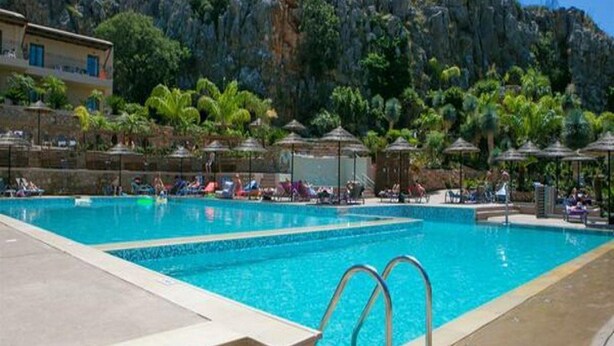 Piscina com espreguiçadeiras e guarda-sóis, cercada por vegetação e um penhasco rochoso.