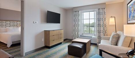 Quarto de hotel com cama, escrivaninha, sofá, televisão e janela com cortinas.