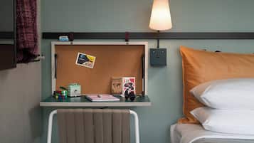 Quarto pequeno e bem organizado, com uma cama, uma escrivaninha e uma luminária.