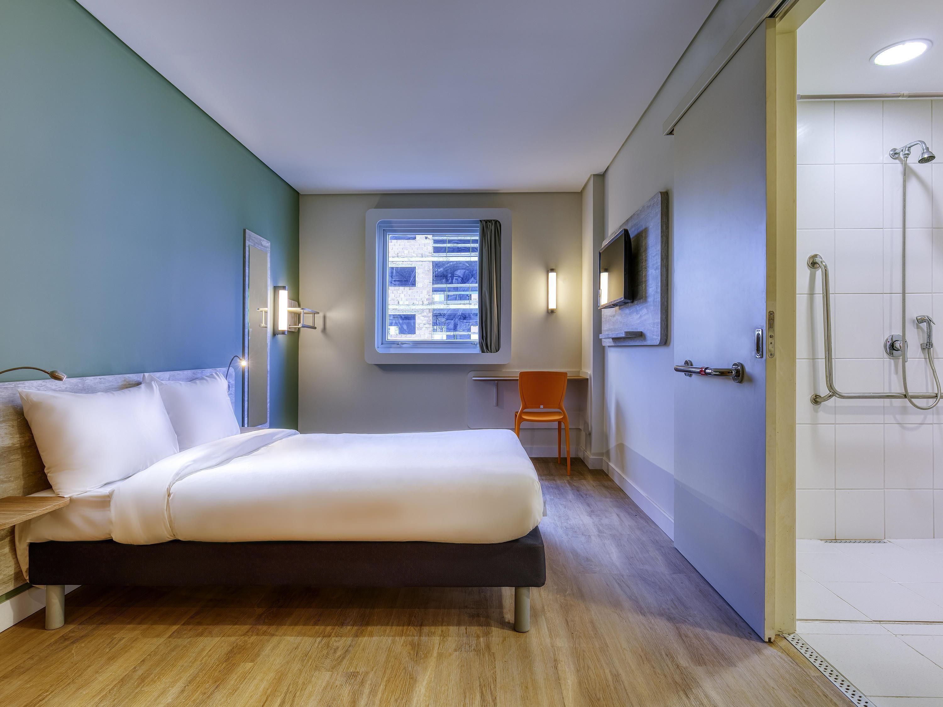Quarto de hotel com cama, escrivaninha, cadeira, televisão e área de chuveiro.
