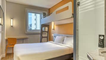 Quarto de hotel com beliche, uma cama de solteiro, janela com vista para edifícios, pequena escrivaninha e banheiro com bancada de granito.