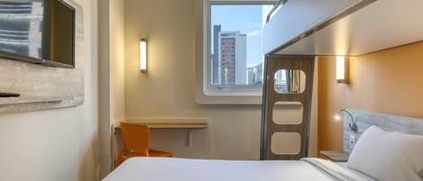 Quarto de hotel com cama, escrivaninha, cadeira, janela com vista para a cidade e espelho fixado na parede.