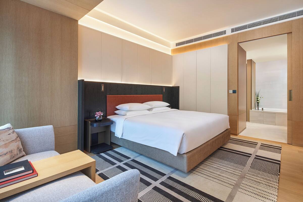 Quarto de hotel moderno com uma cama grande, uma mesinha de cabeceira, um sofá e um banheiro visível através de uma porta aberta.