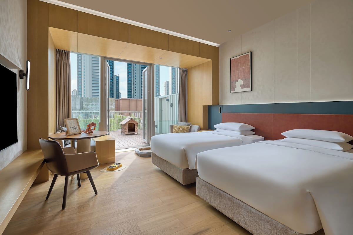 Quarto de hotel com duas camas, uma escrivaninha, uma cadeira e uma janela grande com vista para a cidade.