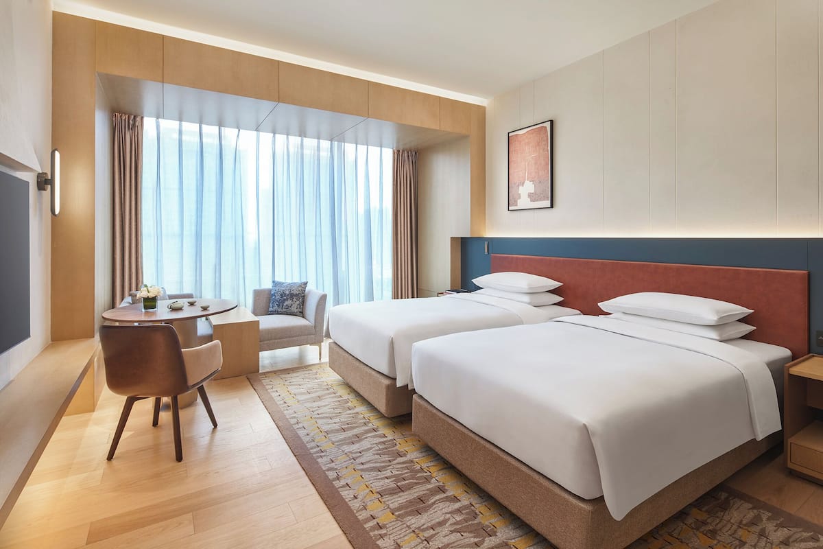 Quarto de hotel com duas camas, uma escrivaninha, uma cadeira e uma janela grande com cortinas.