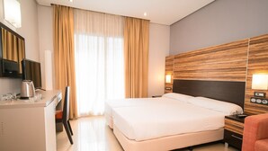 Quarto de hotel com cama, mesa de cabeceira, luminária e escrivaninha com chaleira.