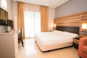 Quarto de hotel com cama, mesa de cabeceira, luminária e escrivaninha com chaleira.