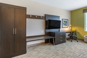 Quarto de hotel moderno com um guarda-roupa espaçoso, uma televisão de tela plana na parede, uma escrivaninha com gavetas, uma cadeira e um quadro na parede.