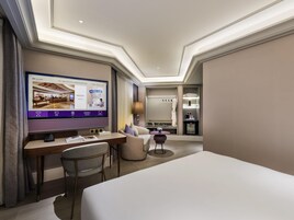 Quarto de hotel com uma televisão de tela grande, uma escrivaninha com cadeira, uma cama, um sofá e uma pequena mesa com abajur.
