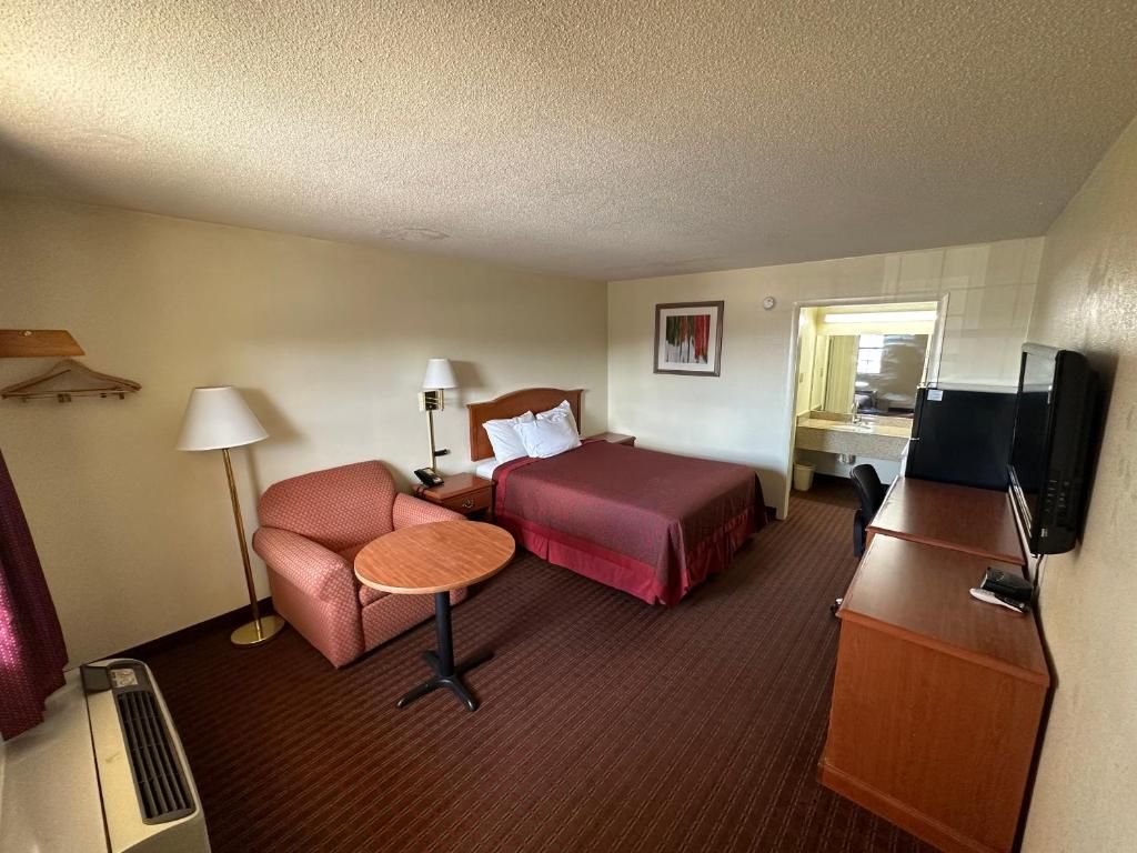 Quarto de hotel com cama, escrivaninha, cadeira, mesa pequena, abajur e televisão.
