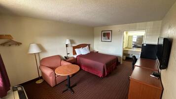 Quarto de hotel com cama, escrivaninha, cadeira, mesa pequena, abajur e televisão.