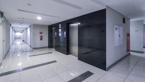 Um corredor comprido e estreito, com piso de azulejos, portas e equipamentos de emergência.