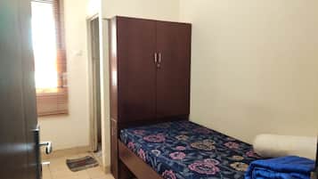 Quarto pequeno com uma cama de solteiro, guarda-roupa de madeira, janela com persianas e um cobertor azul dobrado sobre a cama.