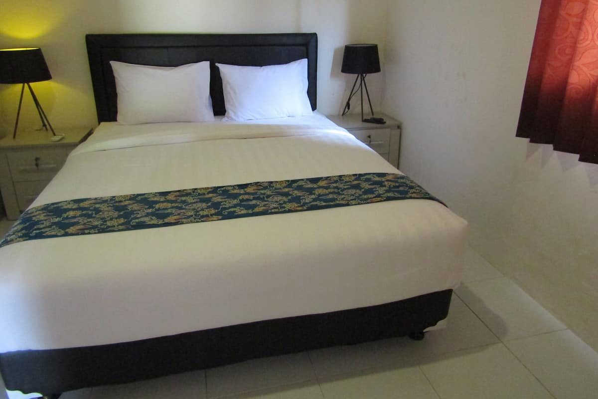 Uma cama bem arrumada com lençóis brancos e um cobre-leito decorativo azul e dourado. Há duas mesas de cabeceira com abajures, e uma cortina vermelha está parcialmente visível à direita.
