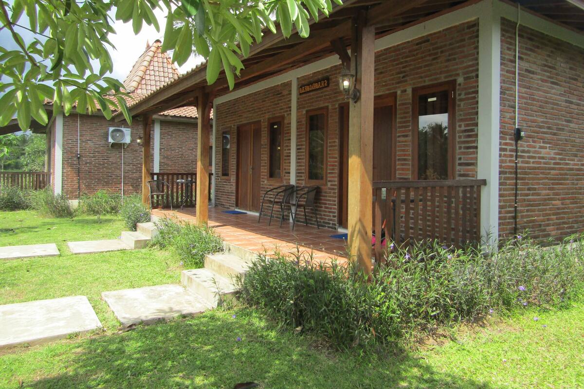 Uma casa de tijolos com varanda, um pequeno jardim e uma placa com a inscrição 'GANGSA SANGGA 2'.