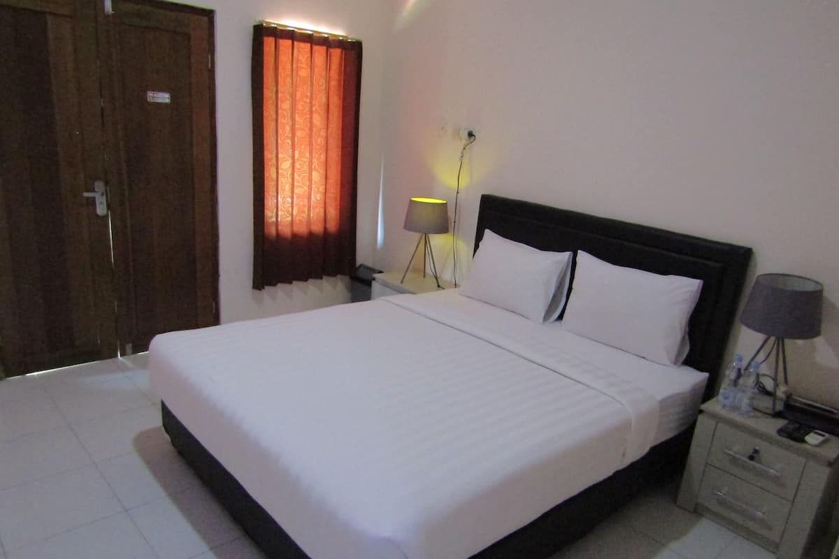 Quarto de hotel padrão com uma cama grande, mesa de cabeceira com abajur, porta de madeira e janela com cortinas.
