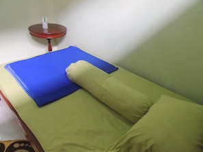 Quarto pequeno com uma cama de solteiro, colchão verde e roupa de cama azul. Há uma pequena mesa redonda com uma garrafa sobre ela.