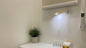 Quarto pequeno e bem iluminado com uma mesa branca, um vaso de planta, uma cadeira e uma estante com garrafas e uma caixa.