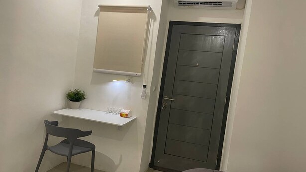 Um quarto pequeno com uma escrivaninha, uma cadeira, uma planta em vaso, uma estante com objetos, uma porta e um aparelho de ar condicionado.