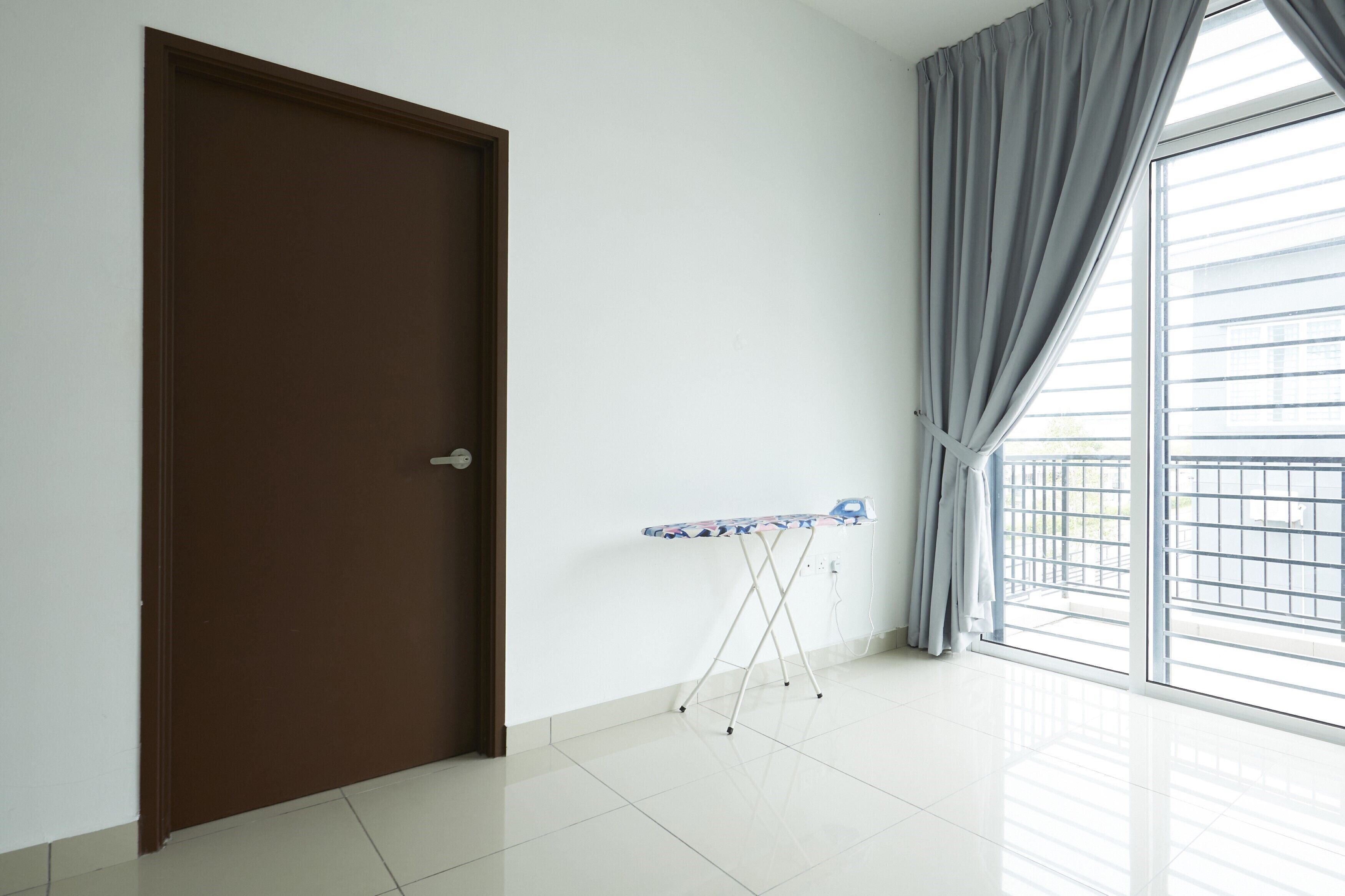 Um quarto com porta fechada, uma tábua de passar pequena e um ferro, e uma janela grande com cortinas.