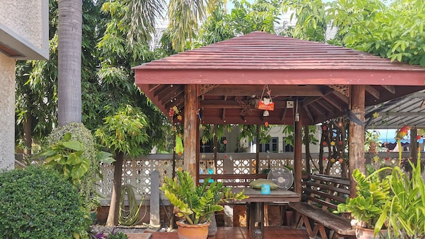 Um gazebo de madeira com telhado de azulejos, cercado por plantas em vasos e um banco.