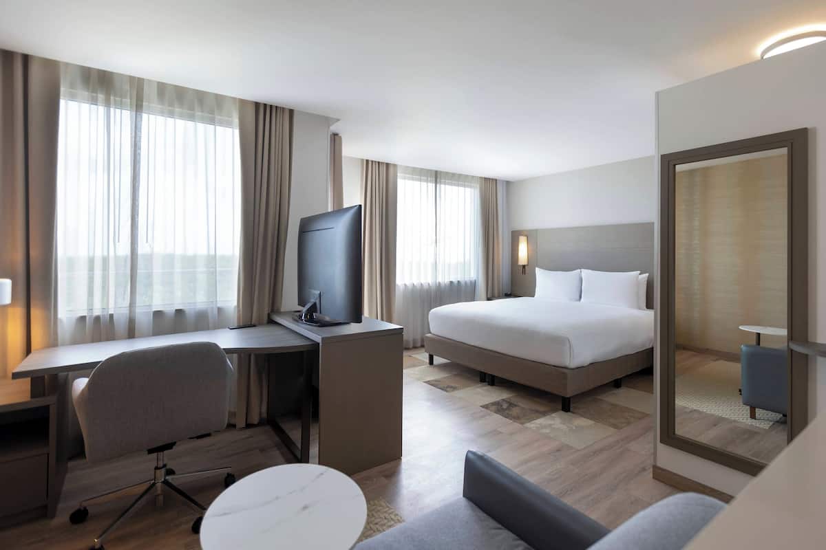 Quarto de hotel com uma cama, uma escrivaninha com cadeira, televisão e uma mesinha.
