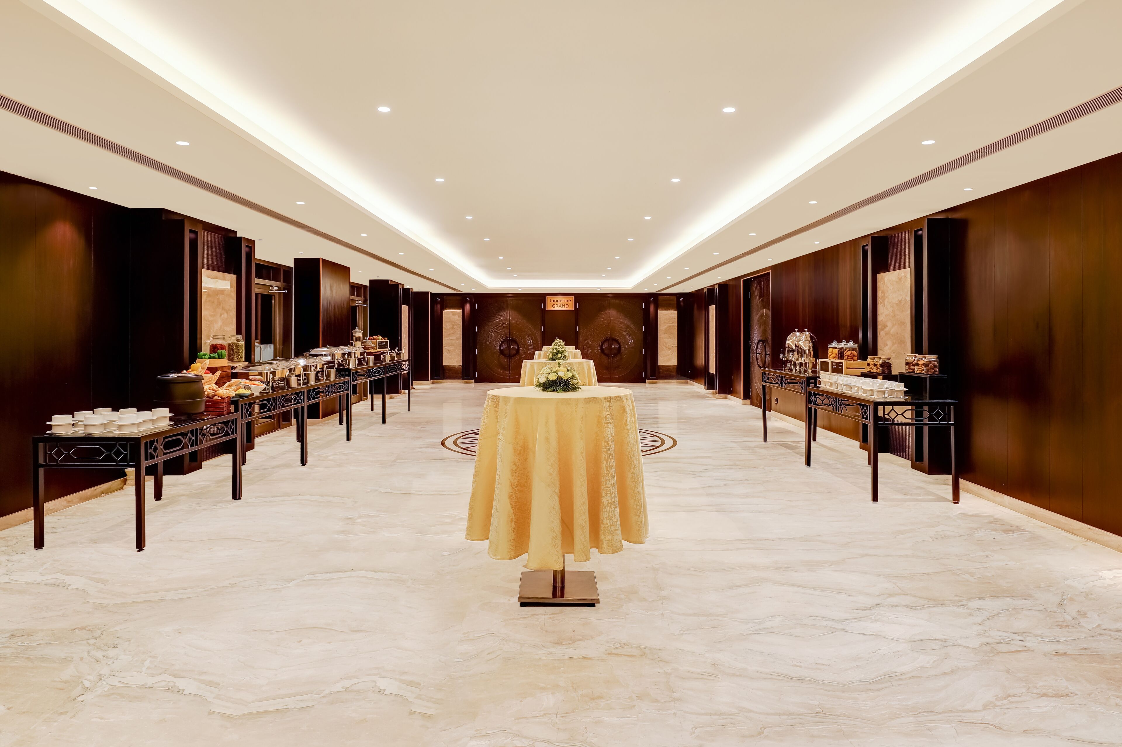 Corredor longo e bem iluminado, com uma mesa central decorada com toalha amarela e arranjo floral, e mesas de buffet laterais com comida e bebida.