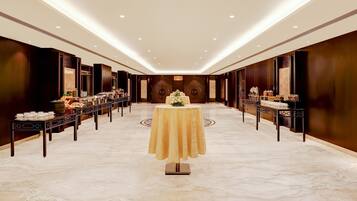 Corredor longo e bem iluminado, com uma mesa central decorada com toalha amarela e arranjo floral, e mesas de buffet laterais com comida e bebida.