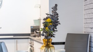 Uma área de refeições moderna com mesa de vidro, duas cadeiras e um vaso com flores.
