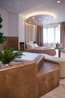 Quarto de hotel moderno com uma cama grande, área de estar, uma mesa pequena com uma garrafa e copos, e uma planta grande.