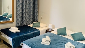 Quarto de hotel com duas camas, cada uma com colcha azul e almofadas brancas. Há um espelho grande na parede, cortina estampada e mesa de cabeceira com abajur.