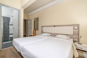 Quarto de hotel com uma cama grande, uma mesa de cabeceira com abajur e área de chuveiro com porta de vidro.