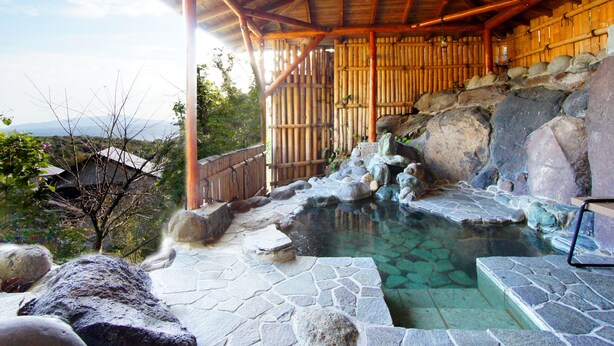 Uma área de águas termais com degraus de pedra que levam a uma piscina, cercada por pedras e uma cerca de bambu.
