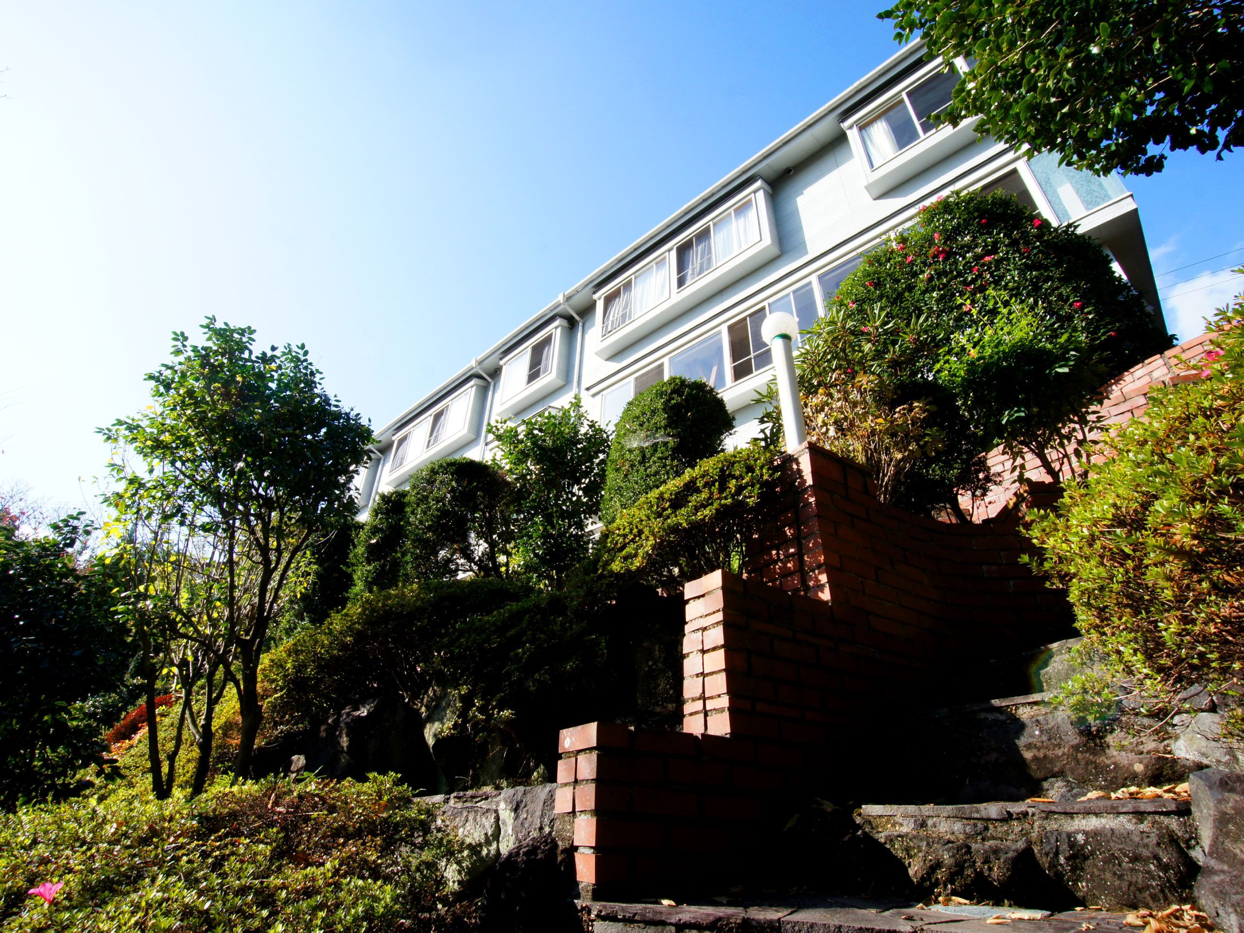 Um edifício com vários andares, base em tijolo e seções superiores brancas, rodeado por vegetação exuberante e árvores.