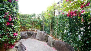 Um jardim com caminho de pedra, rodeado por vegetação exuberante e flores cor-de-rosa.