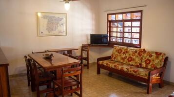 Uma sala de jantar com mesa e cadeiras de madeira, um sofá com estofamento floral, uma televisão sobre um suporte e um ventilador de teto.