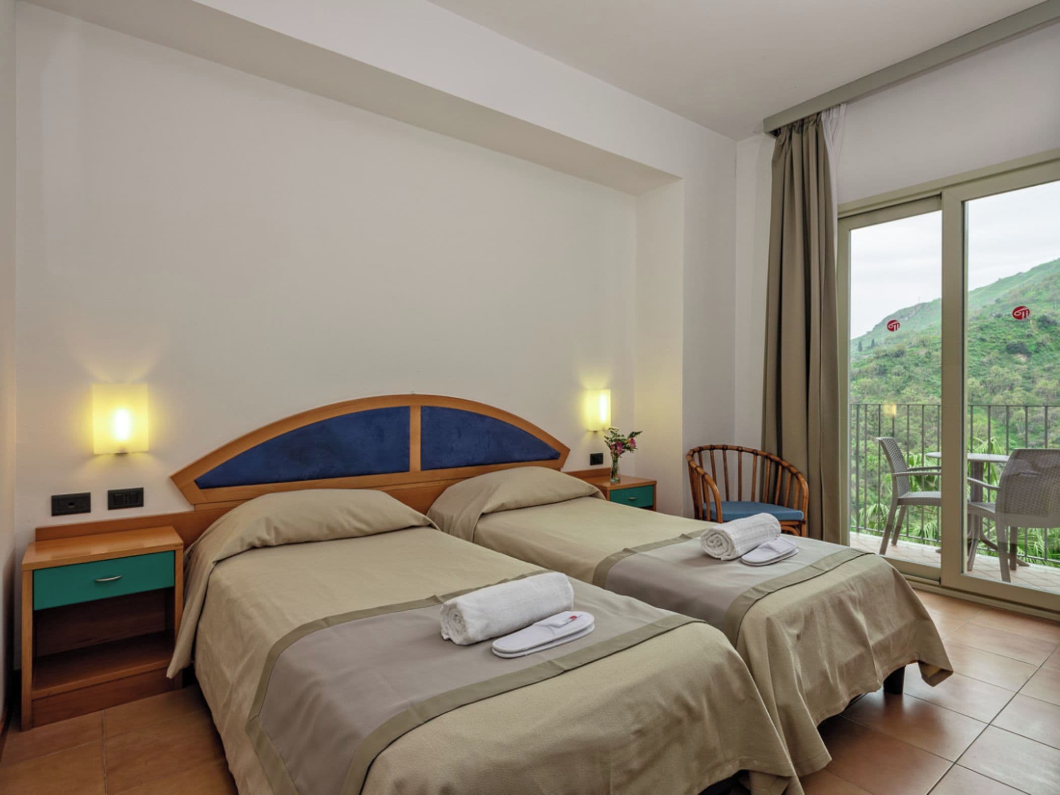 Quarto de hotel com duas camas, uma varanda e vista para colinas, e uma cadeira.